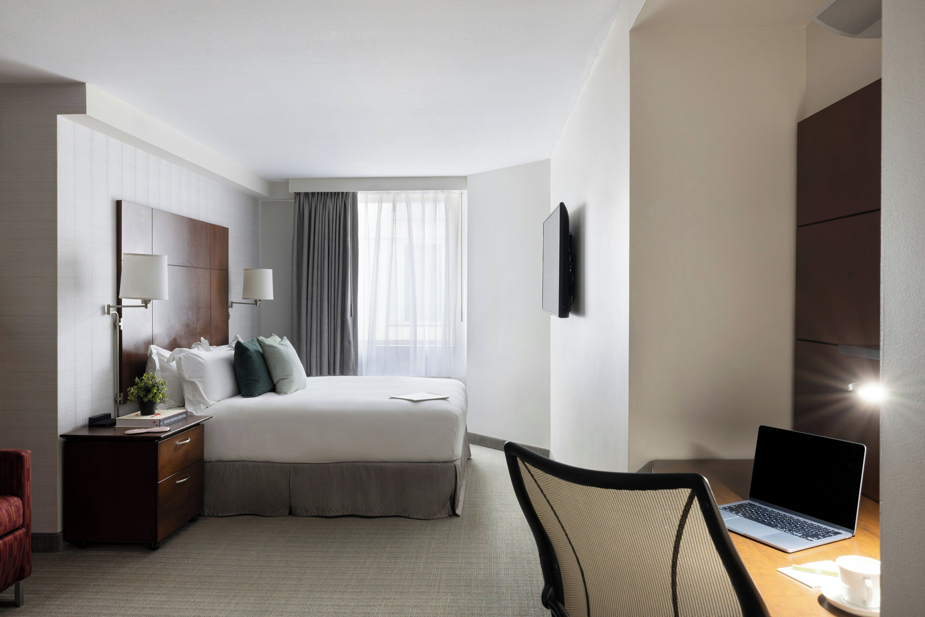 Quarto de hotel com cama, escrivaninha com laptop, cadeira, mesa de cabeceira com abajur e televisão na parede.