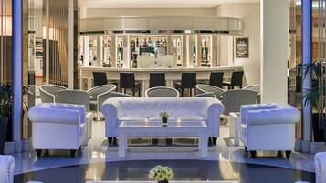 Uma área de estar moderna com um sofá branco central, duas poltronas, uma mesa de centro de vidro com um vaso de flores e um bar com diversos frascos.