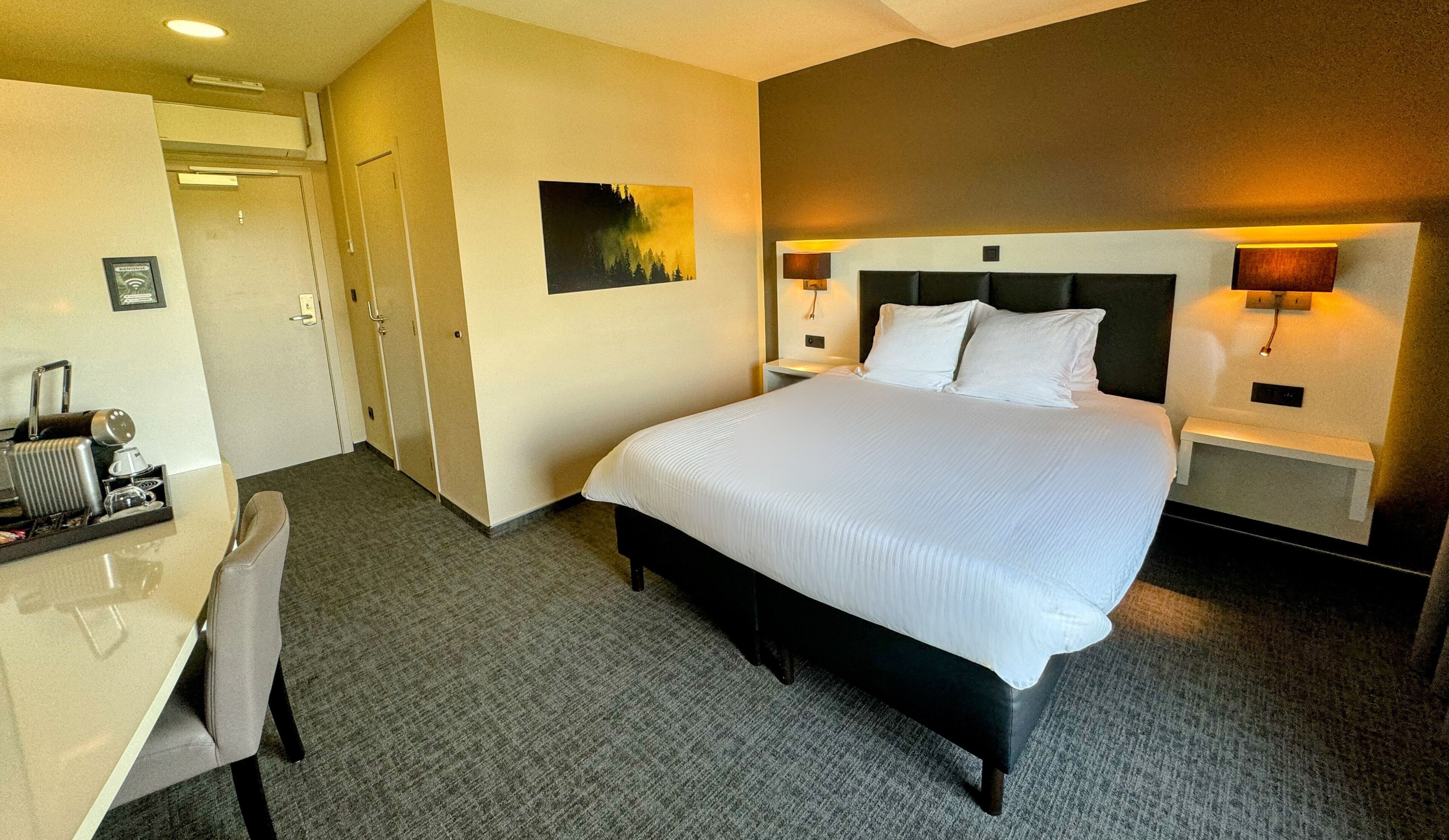 Quarto de hotel com uma cama grande, mesas de cabeceira com luminárias, uma escrivaninha com uma cadeira e uma porta que dá para outro aposento.