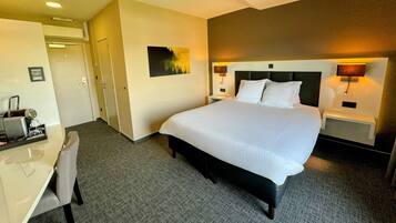Quarto de hotel com uma cama grande, mesas de cabeceira com luminárias, uma escrivaninha com uma cadeira e uma porta que dá para outro aposento.