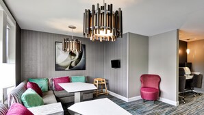 Quarto de hotel moderno com sofá cinzento, almofadas coloridas, mesa branca, cadeira vermelha, televisão e tapete estampado.