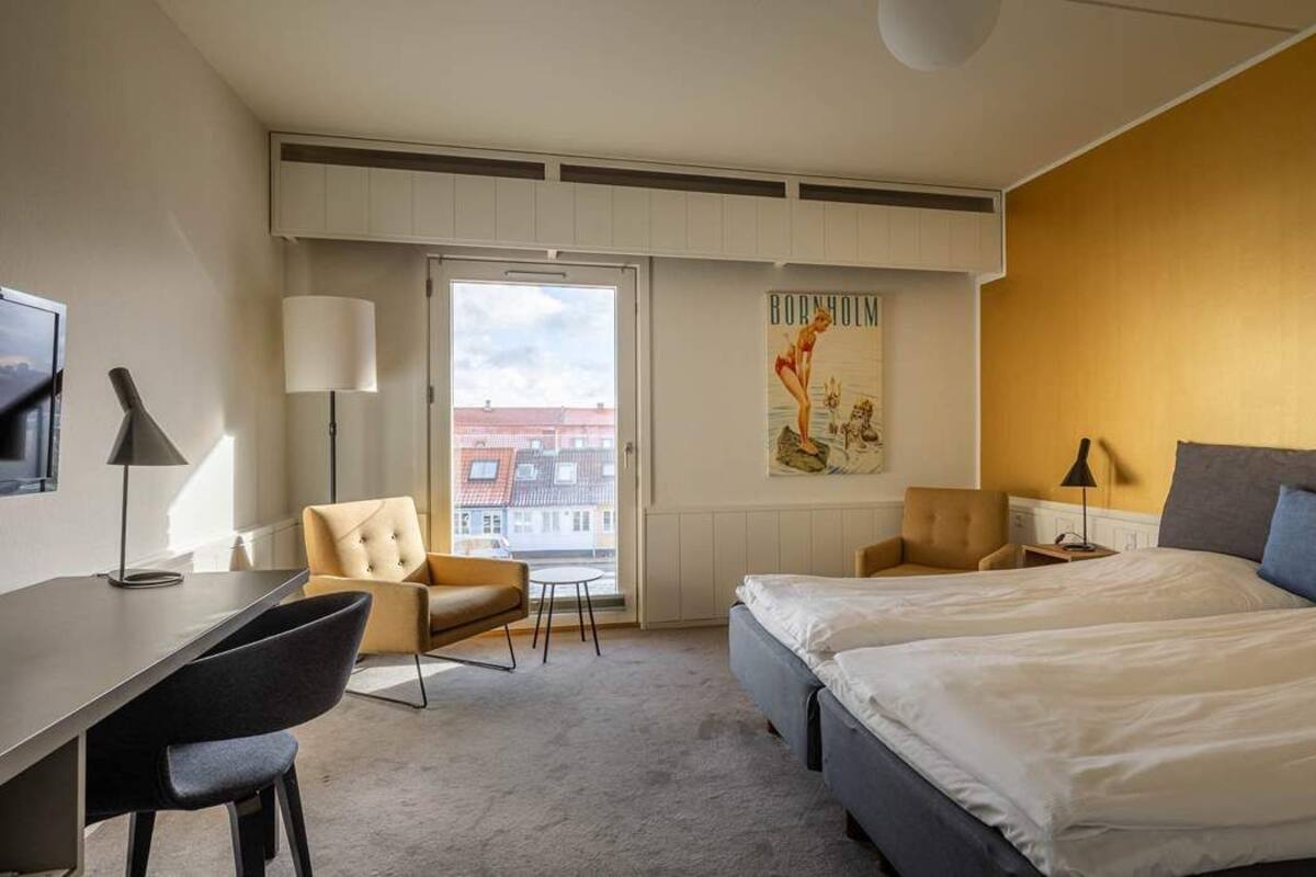 Quarto de hotel com duas camas, uma escrivaninha, uma cadeira, um abajur e um cartaz na parede.