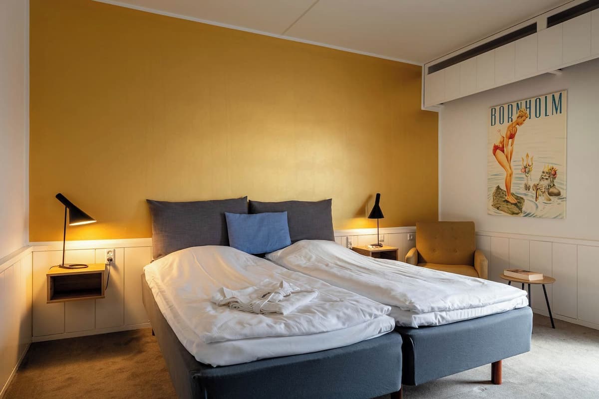 Uma cama de casal com lençóis brancos, duas mesas de cabeceira com abajures, uma cadeira e uma parede com um pôster.