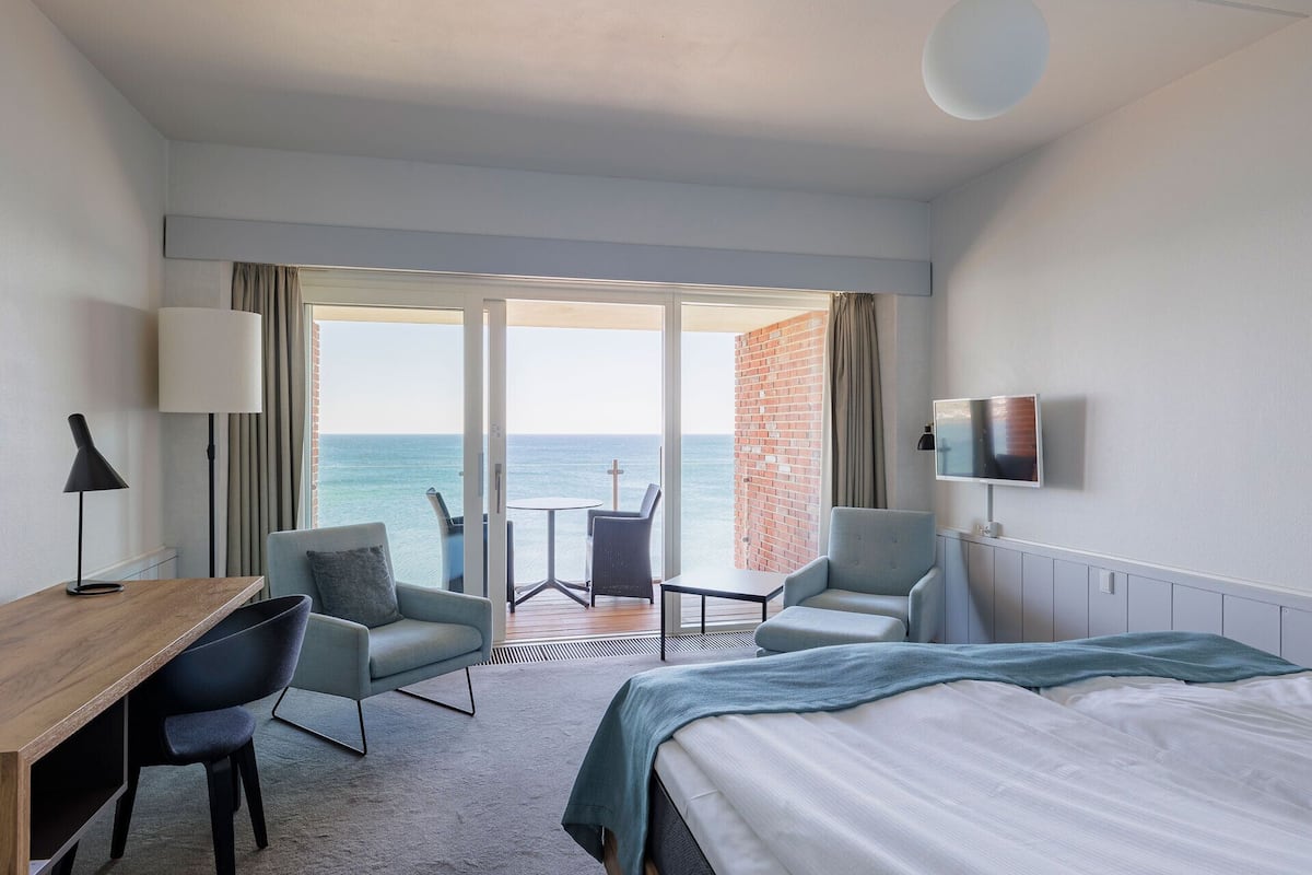 Quarto de hotel com cama, escrivaninha, duas cadeiras, televisão e varanda vista para o mar.