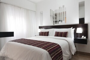 Quarto de hotel com uma cama grande, roupa de cama branca, manta listrada, mesa de cabeceira com abajur e um espelho na parede.