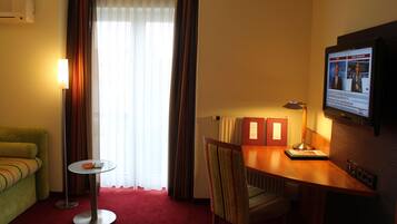 Quarto de hotel com sofá verde, mesa redonda pequena, escrivaninha de madeira com cadeira, televisão e uma grande janela com cortinas.