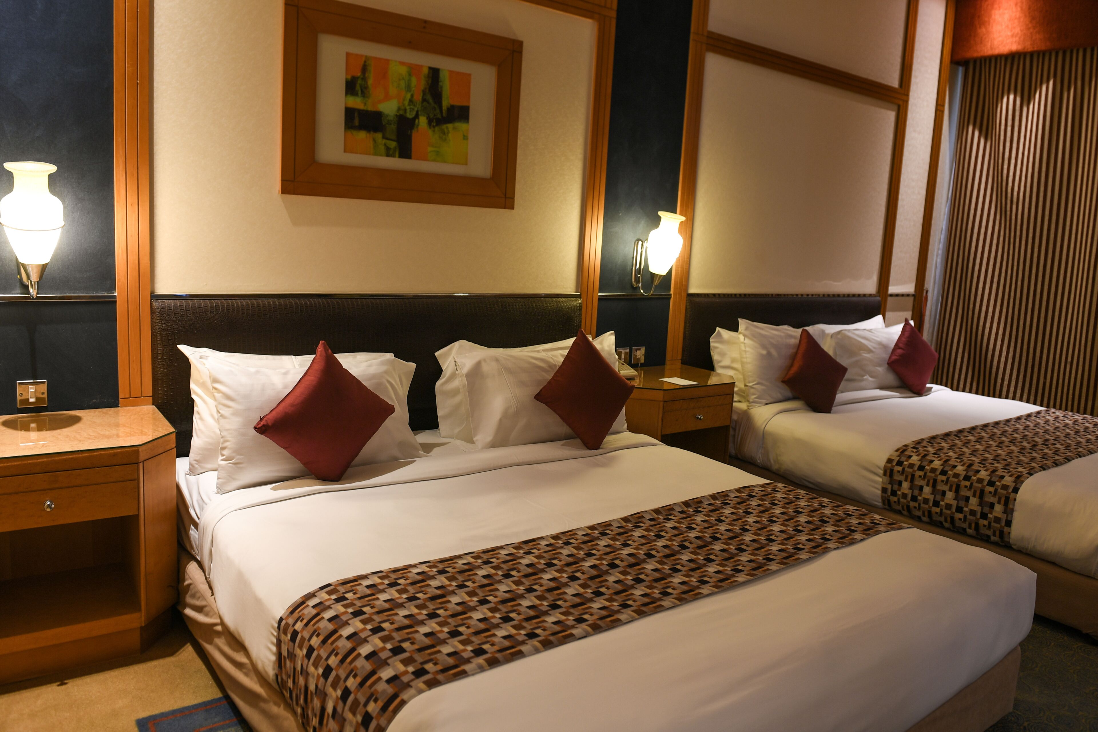 Quarto de hotel com duas camas, cada uma com roupa de cama branca e colchas estampadas. Há duas mesas de cabeceira com abajures, um criado-mudo e um quadro emoldurado na parede.