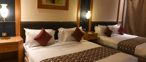 Quarto de hotel com duas camas, cada uma com roupa de cama branca e colchas estampadas. Há duas mesas de cabeceira com abajures, um criado-mudo e um quadro emoldurado na parede.