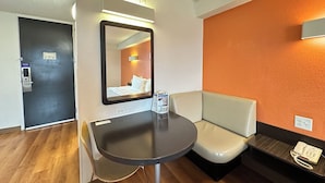 Quarto de hotel com piso de madeira, uma pequena mesa com cadeira, um sofá, um espelho e um telefone em uma prateleira.
