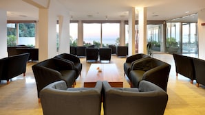 Um hall amplo com uma mesa de centro, sofás pretos e grandes janelas com vista para o mar.