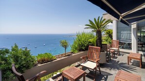 Varanda com móveis de madeira, uma palmeira e vista para o oceano com barcos.