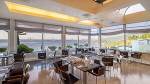 Ampla sala de jantar com grandes janelas que proporcionam vista para o mar e uma ponte. Mesas montadas com cadeiras e utensílios para refeições.