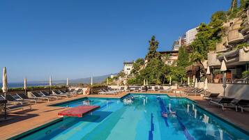 Uma área de piscina com espreguiçadeiras e guarda-sóis, cercada por edifícios e árvores.