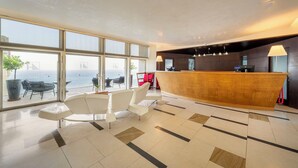 Uma área de recepção de hotel com um balcão de madeira, cadeiras brancas e grandes janelas com vista para o mar.