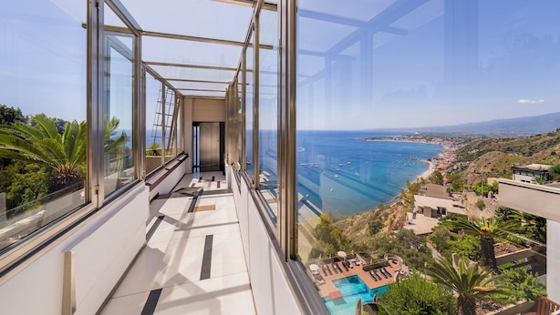 Um edifício alto com uma galeria de vidro que dá para uma varanda com piscina e vista para o oceano.