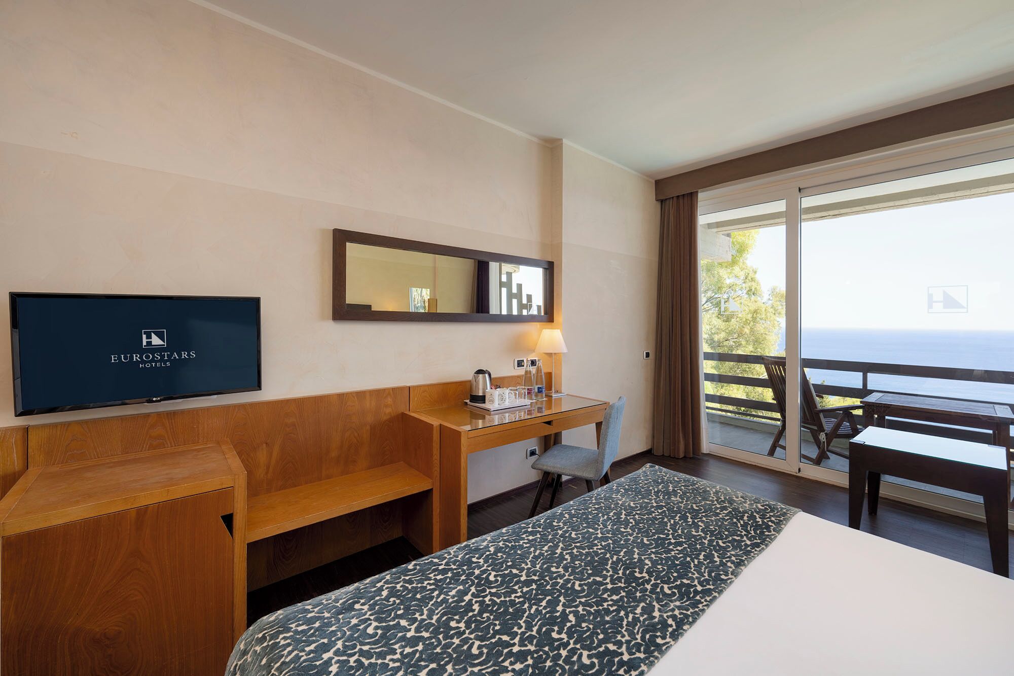 Quarto de hotel com cama, escrivaninha, cadeira, televisão, espelho e varanda com vista para o mar.