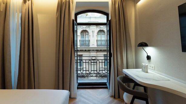 Quarto de hotel com cama, escrivaninha e vista para um edifício através de uma porta de varanda.