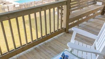 Um deck de madeira com cadeiras brancas e uma pequena mesa, com vista para uma piscina e praia.