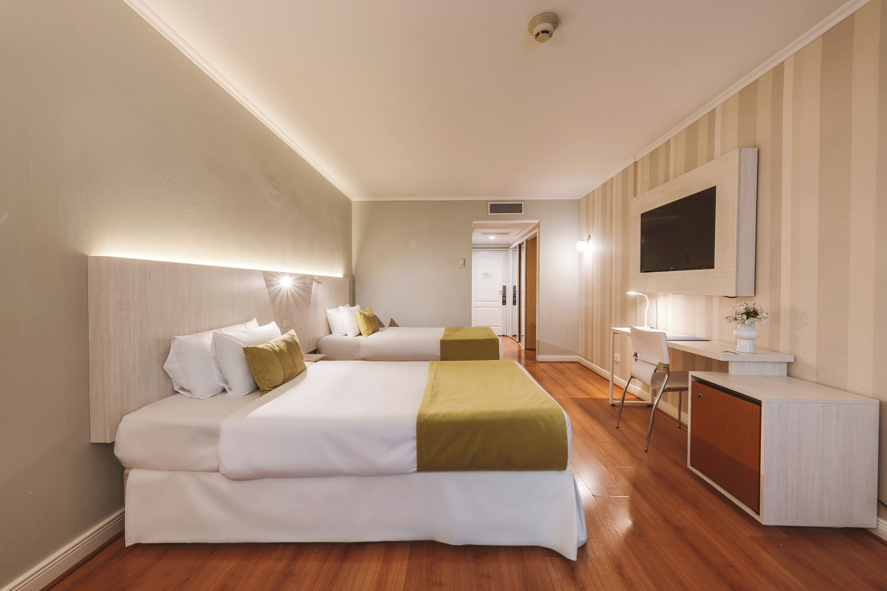 Quarto de hotel com duas camas, uma escrivaninha, uma cadeira, uma televisão e uma porta para o banheiro.