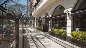 Uma calçada com mesas ao ar livre, um edifício com janelas em arco e um céu claro.