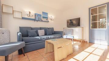 Uma sala de estar moderna com um sofá cinzento, uma mesa de centro de madeira e uma TV sobre um suporte. Há luminárias fixadas na parede e quadros emoldurados acima do sofá.