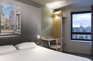 Quarto de hotel com cama, escrivaninha, cadeira, janela com vista para edifícios e luminária na parede.