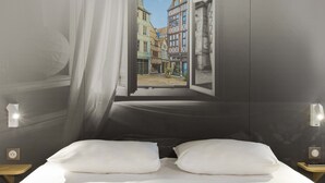 Cama com almofadas brancas e vista para edifícios coloridos pela janela.