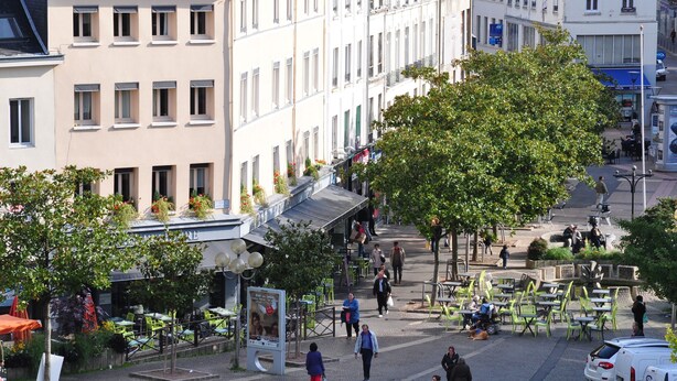 Vista da rua com edifícios, mesas ao ar livre e pessoas passando.