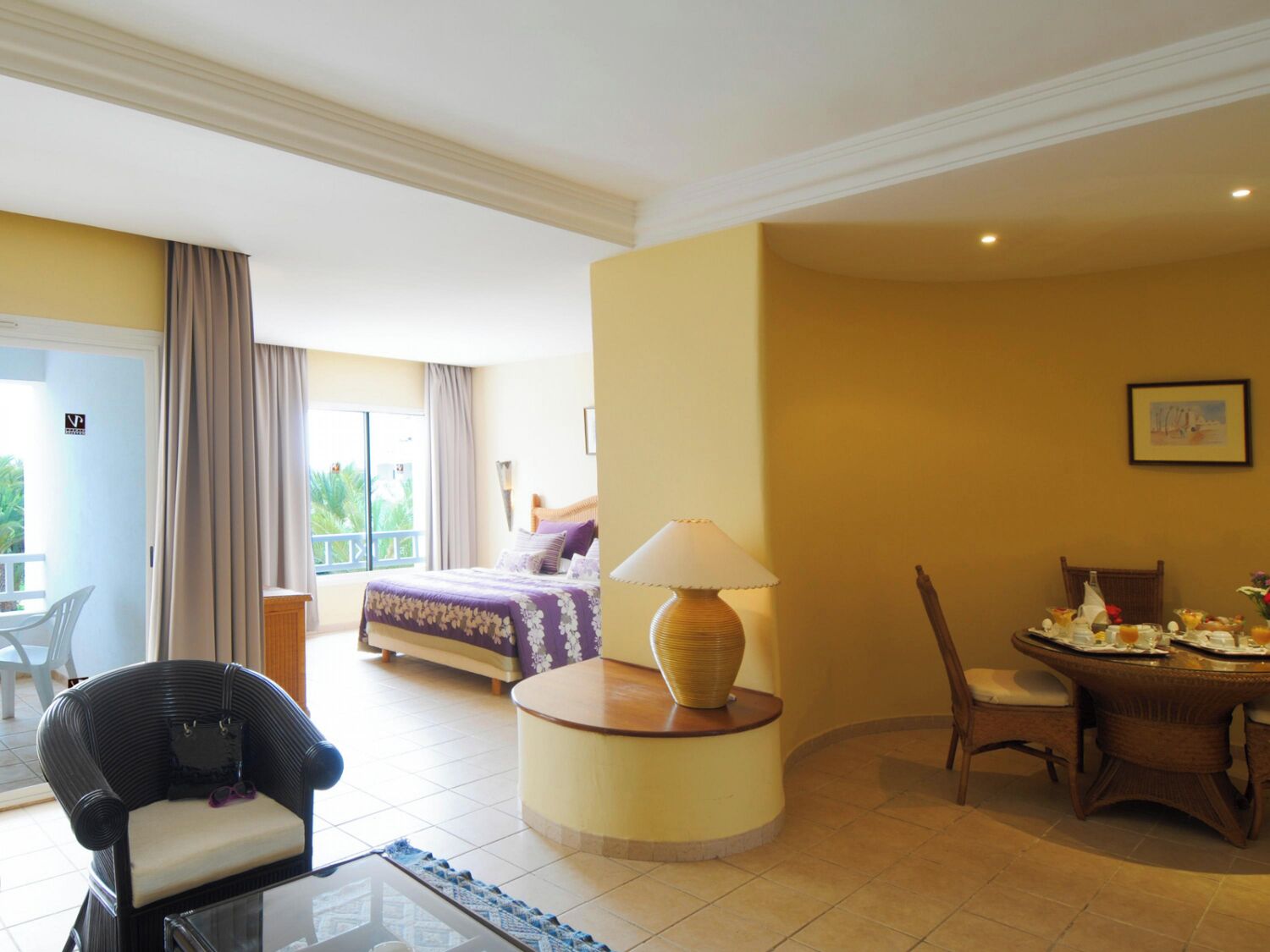 Quarto de hotel com cama, uma mesa de jantar com cadeiras, uma poltrona com uma bolsa, uma luminária e vista para o exterior.