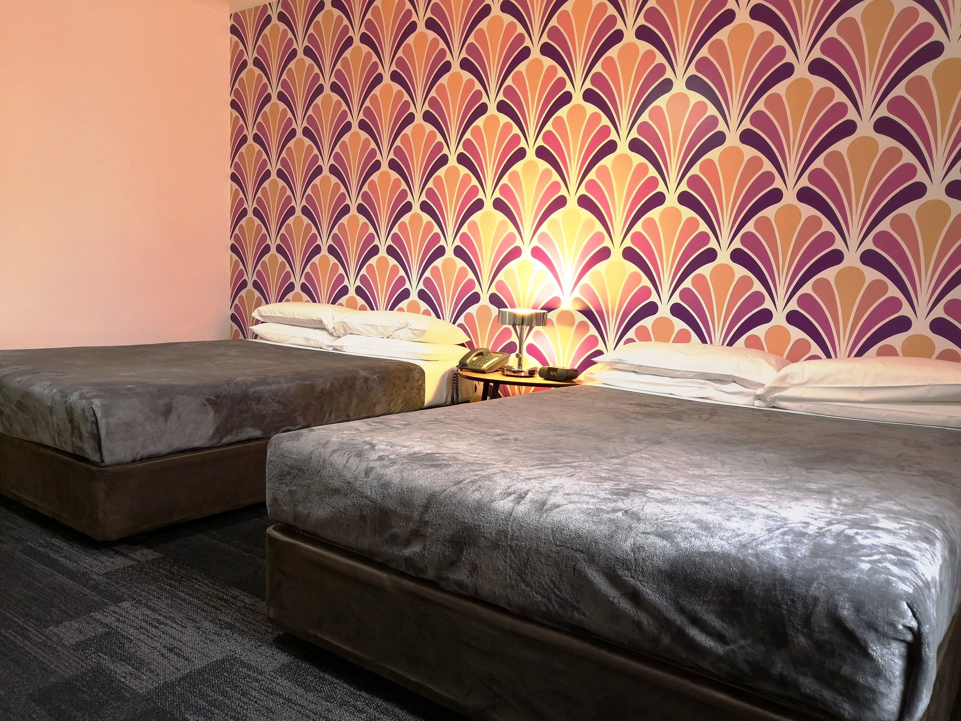 Quarto de hotel com duas camas, uma mesa de cabeceira com abajur e papel de parede estampado.