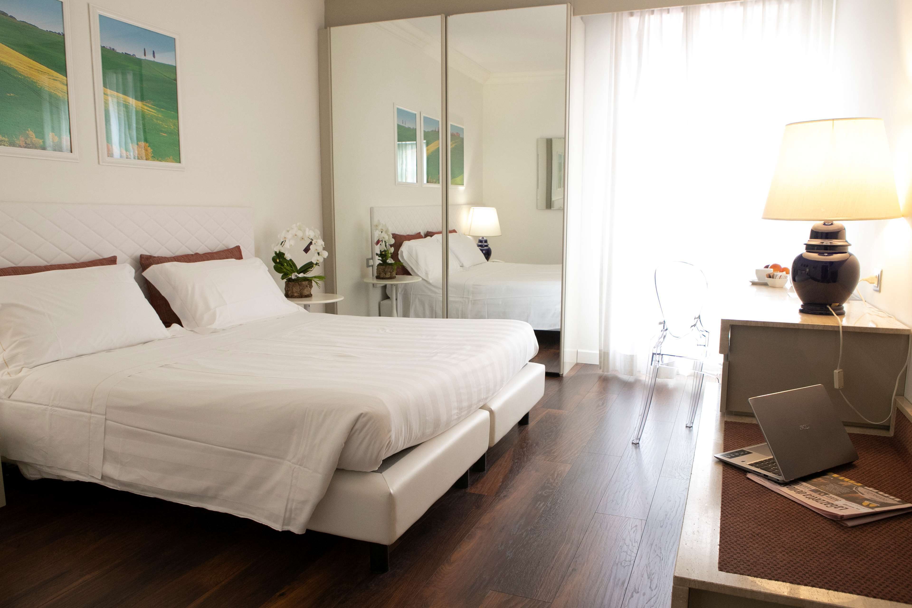 Quarto de hotel com uma cama grande, uma escrivaninha com um laptop, uma luminária e um espelho.