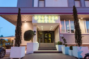 Entrada do Hotel Etrusco com uma placa iluminada em destaque, um conjunto de degraus levando à entrada e fachada de um edifício moderno.