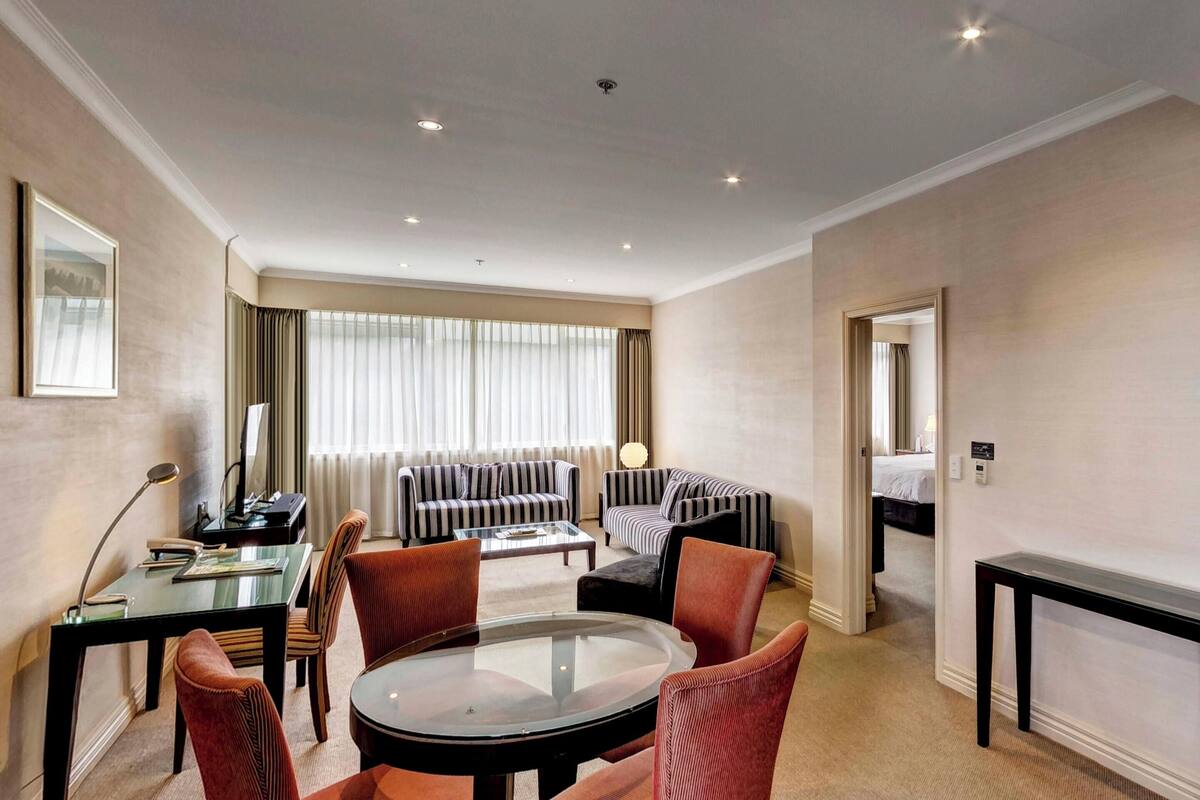 Quarto de hotel moderno com área de estar, mesa de jantar com cadeiras, sofá e uma pequena mesa com luminária.