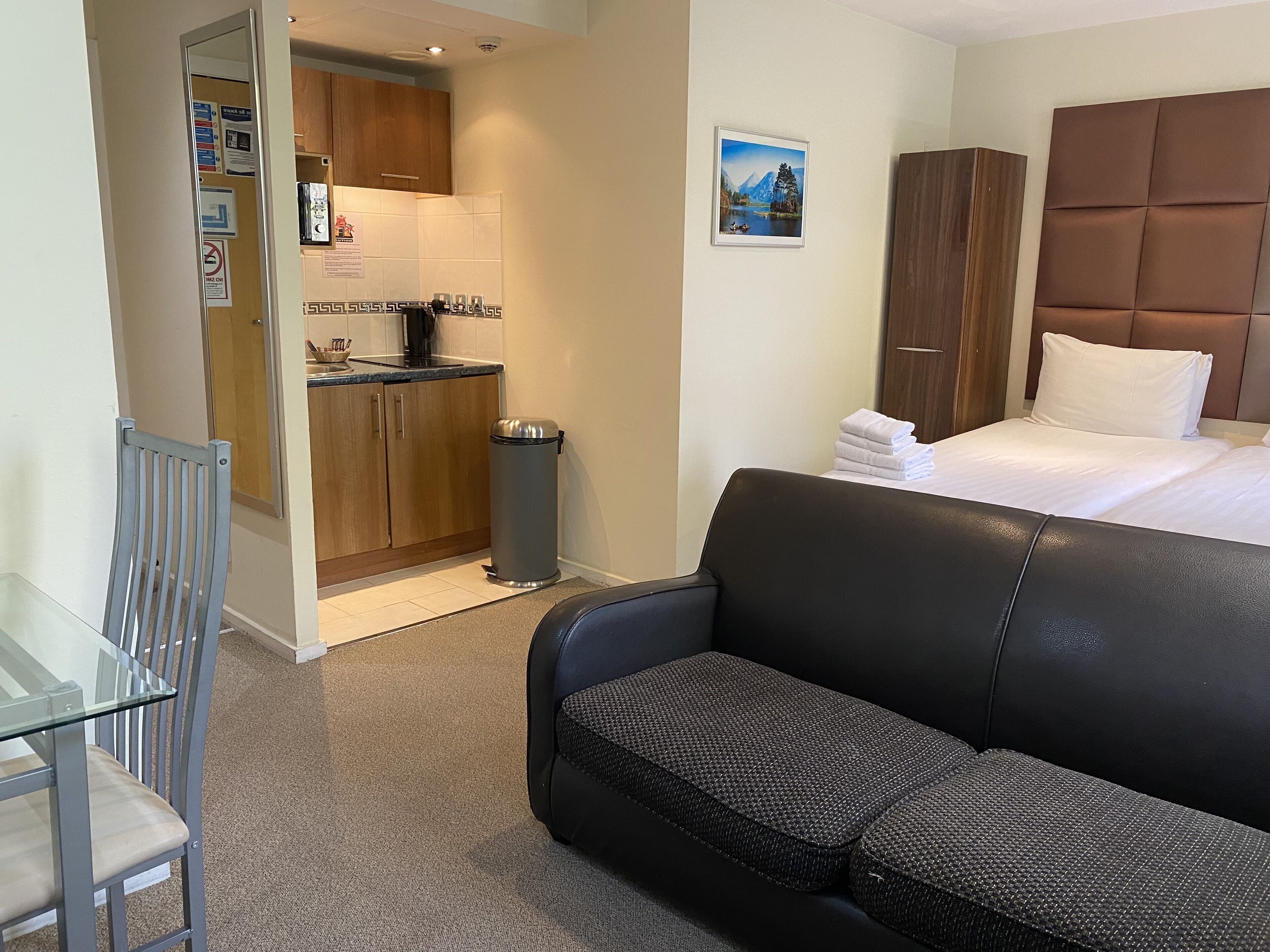 Quarto de hotel com cama, sofá de couro preto, pequena mesa de jantar com cadeira, área de cozinha e lixeira.