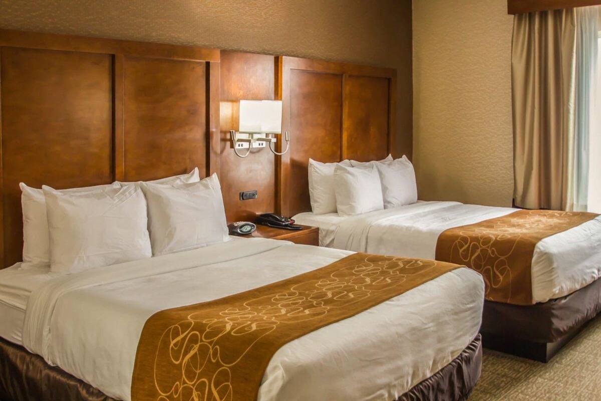 Quarto de hotel com duas camas, cabeceira de madeira, mesa de cabeceira com abajur e janela com cortinas.