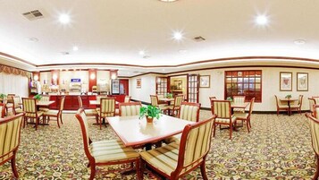 Uma área de refeições de hotel com mesas e cadeiras redondas, um balcão de recepção e um tapete estampado.