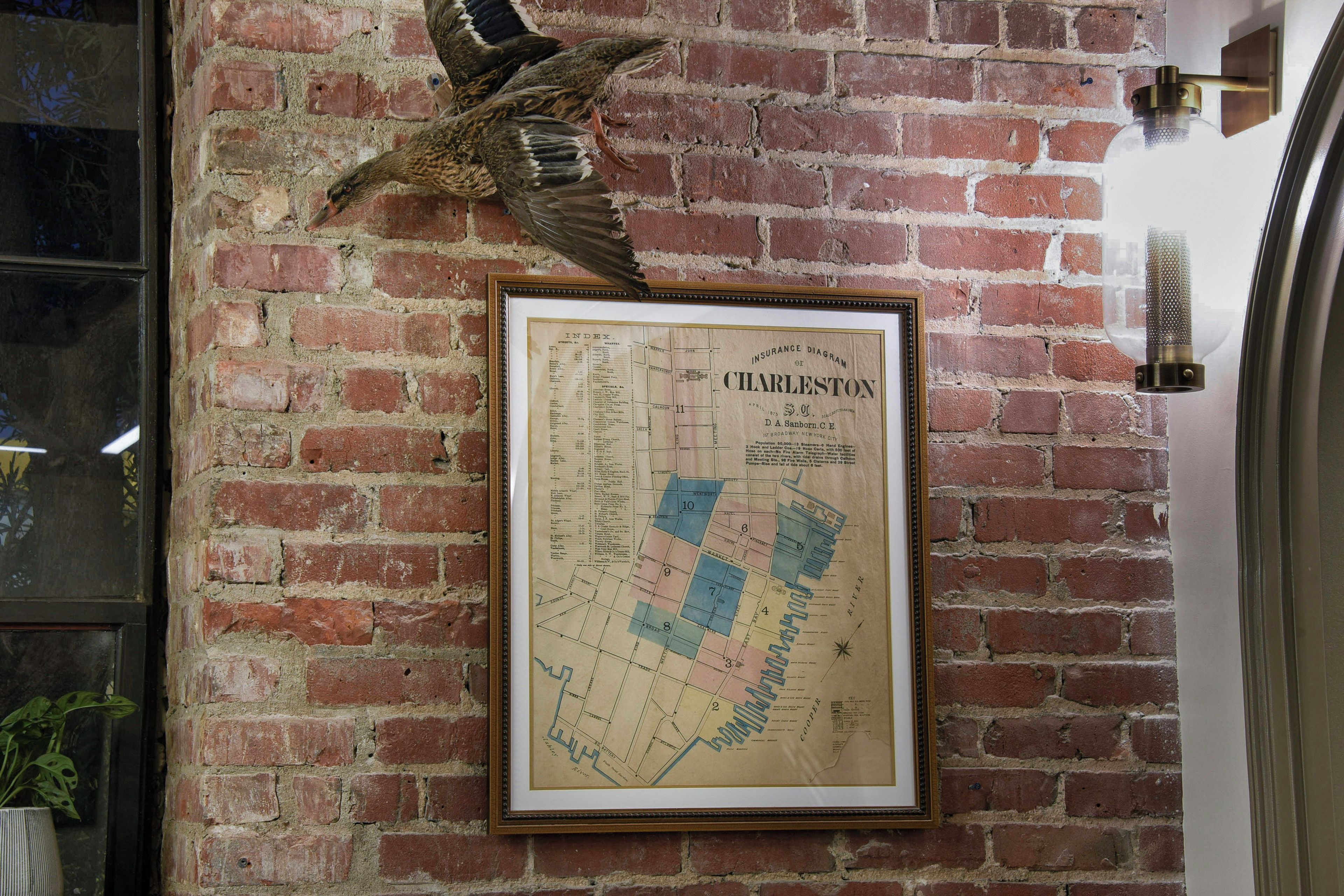 Mapa histórico de Charleston, Carolina do Sul, emoldurado e fixado numa parede de tijolos, com um candeeiro de latão em cima.