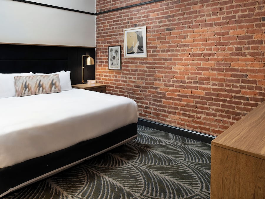 Quarto de hotel com parede de tijolos, uma cama com roupa de cama branca, mesa de cabeceira com abajur e uma pequena planta na escrivaninha.