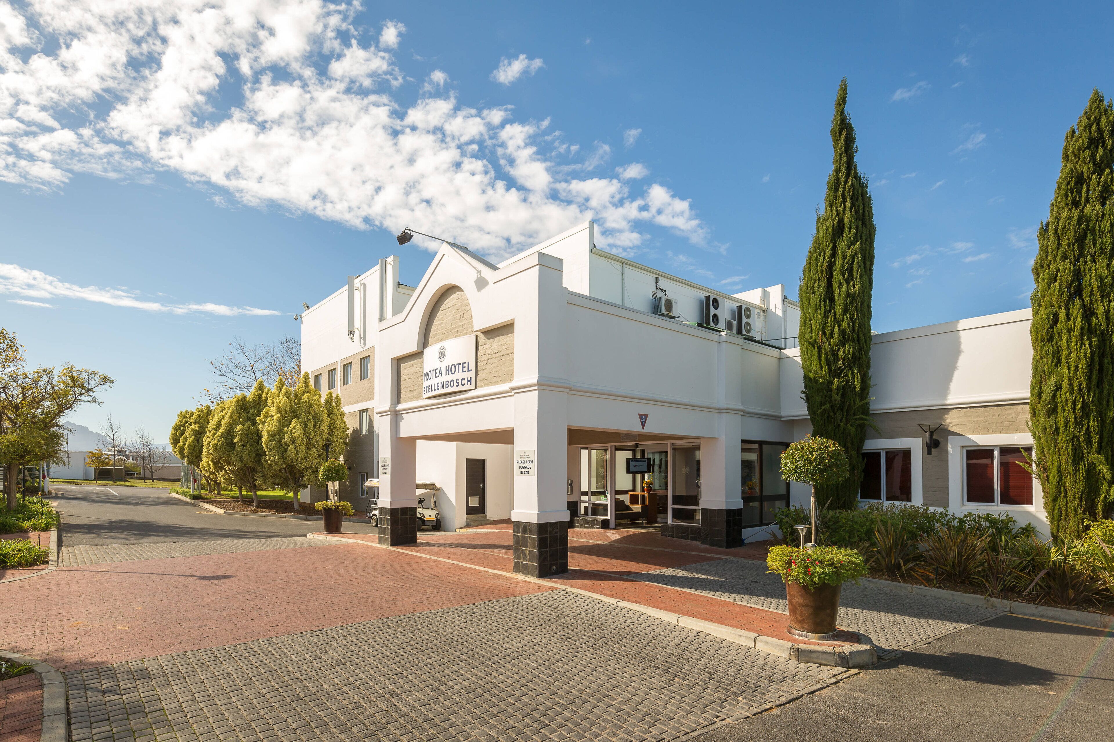 Um hotel com design arquitetônico moderno, destacando uma entrada imponente, área de estacionamento e um jardim bem cuidado com vasos de plantas.