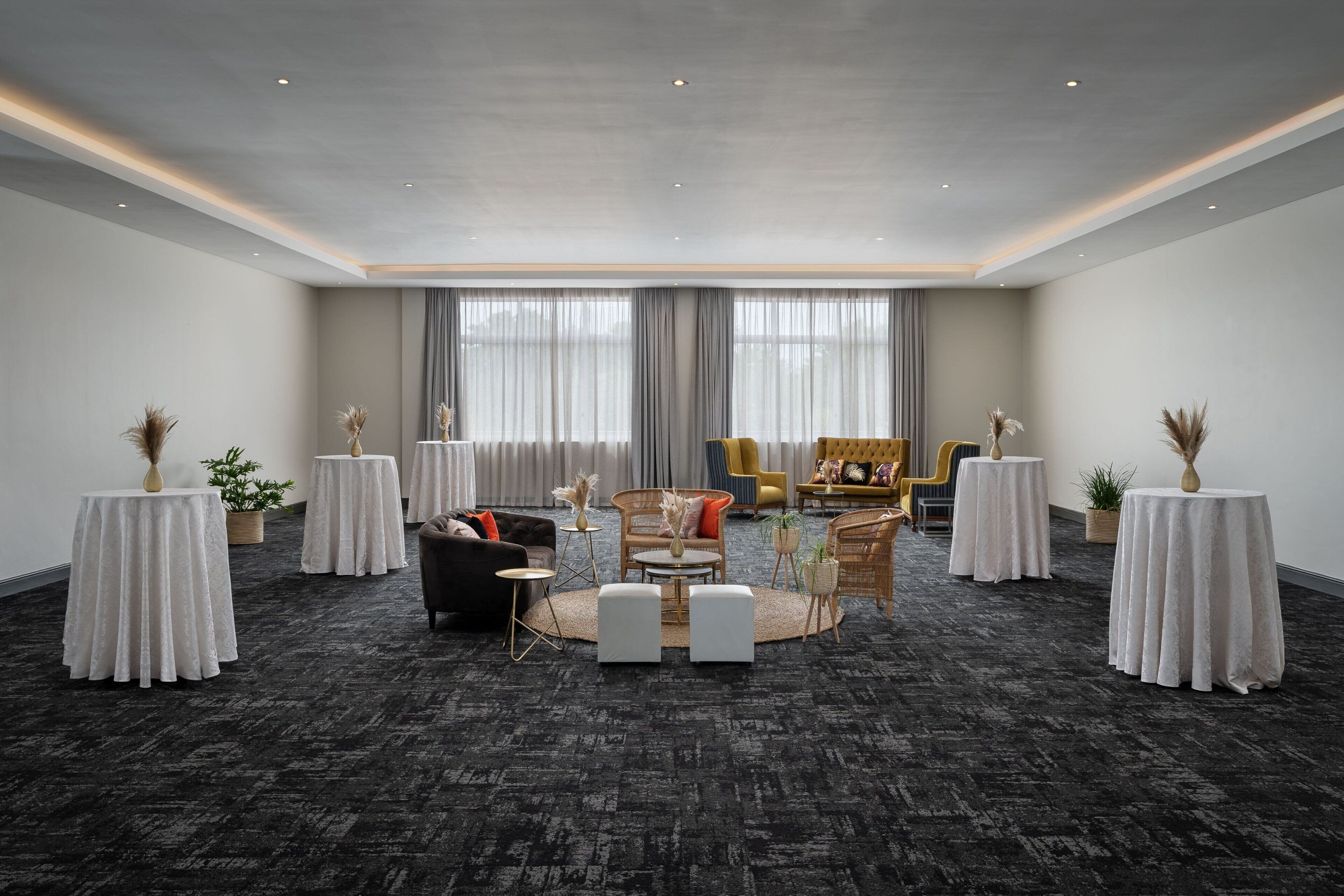 Quarto de hotel moderno com piso de madeira escura, um sofá preto, uma poltrona amarela e uma mesa de centro redonda. Há duas mesinhas de cabeceira com toalhas brancas e plantas decorativas. O quarto possui grandes janelas com cortinas leves e vista para a cidade.