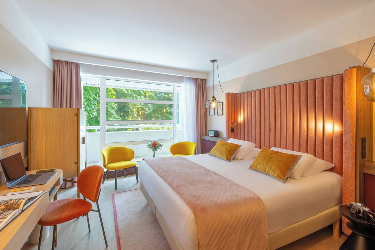 Quarto de hotel moderno com uma cama grande, uma escrivaninha com um laptop, duas cadeiras, uma TV e uma janela com vista para áreas verdes.