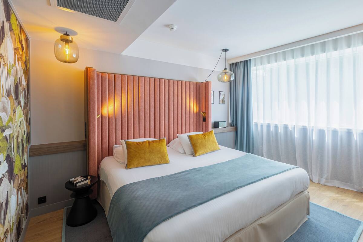 Quarto de hotel moderno com uma cama grande, cabeceira com ripas verticais, duas luminárias de cabeceira, uma mesinha com telefone e um livro, e uma janela com cortinas translúcidas.