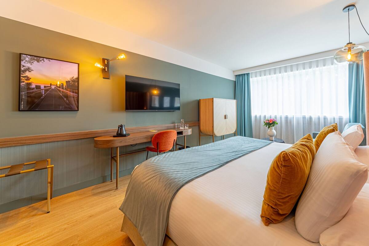 Quarto de hotel com cama, escrivaninha, cadeira, televisão e janela com cortinas.