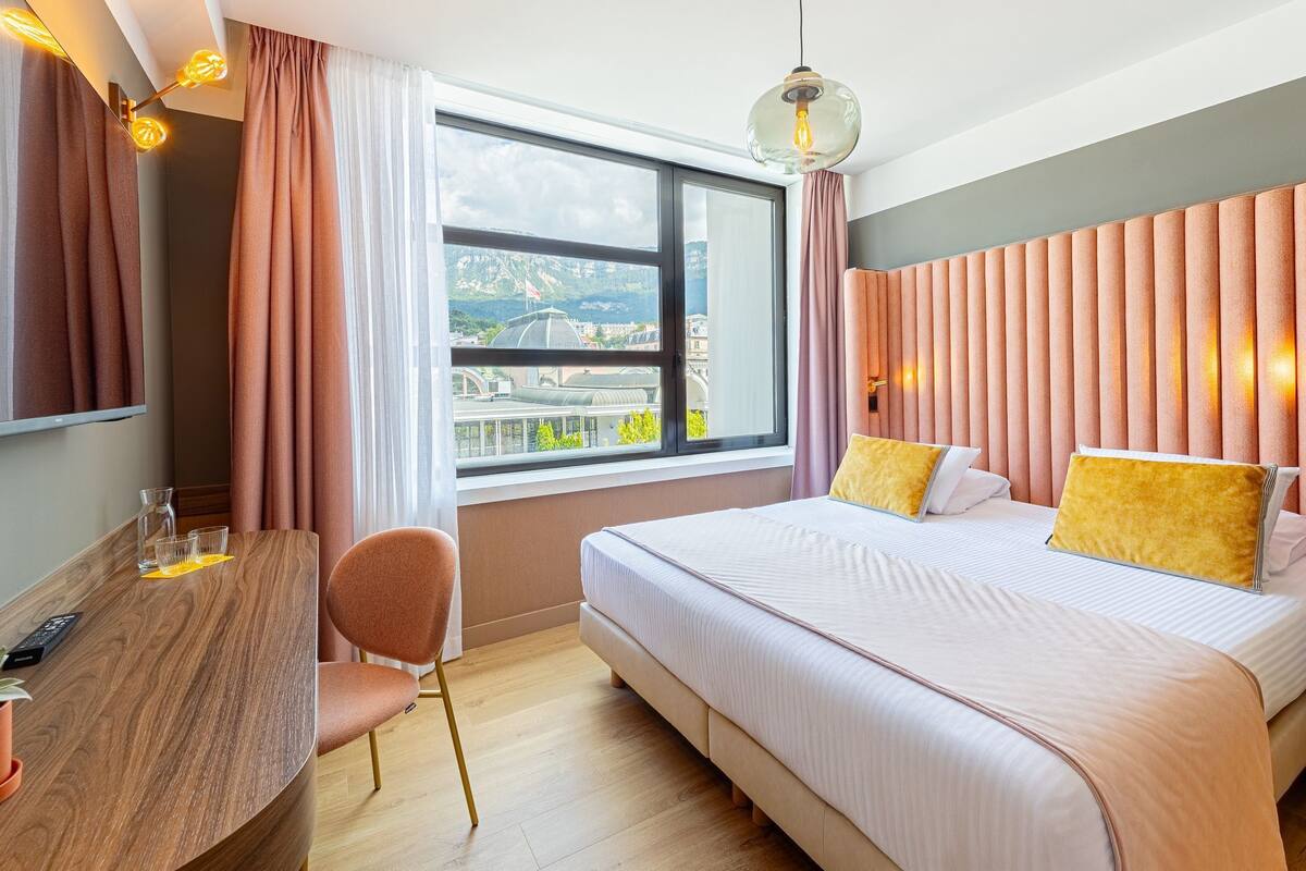 Quarto de hotel moderno com uma cama grande, uma escrivaninha de madeira, uma cadeira e vista para a cidade pela janela.