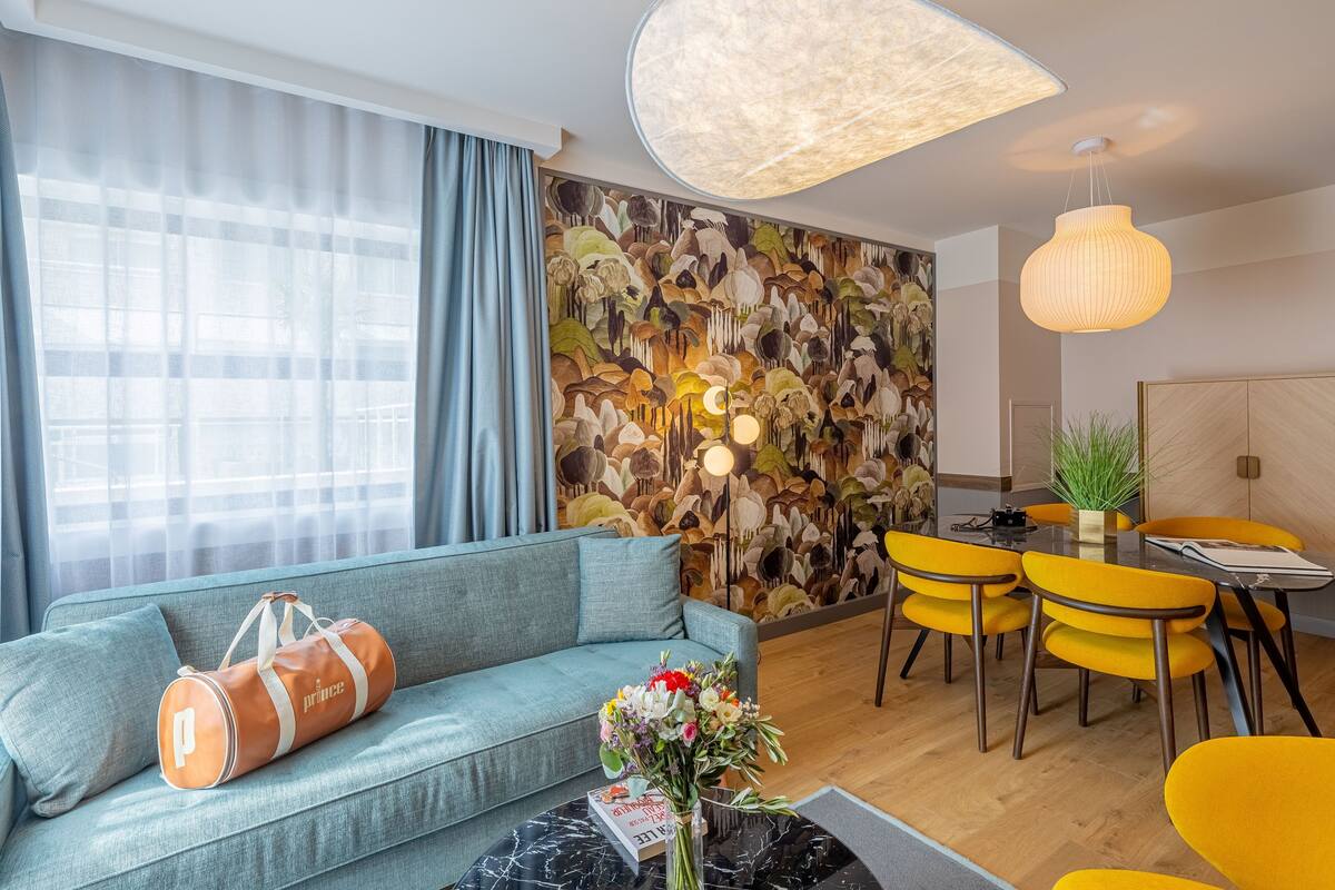 Uma sala de estar moderna com um sofá, uma mesa de jantar com cadeiras amarelas e uma parede com um mural de tema natural.