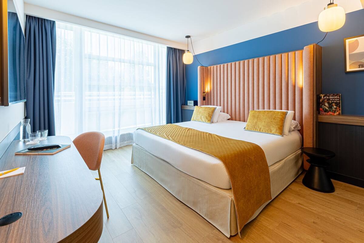 Quarto de hotel com uma cama grande, uma escrivaninha com cadeira, uma mesinha de cabeceira, uma janela com cortinas e uma prateleira na parede com um livro.