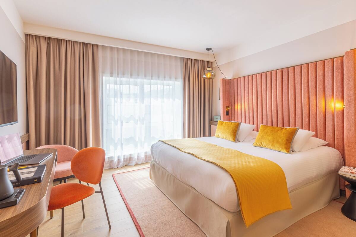 Quarto de hotel com uma cama grande, uma escrivaninha com um laptop, uma cadeira e uma televisão.