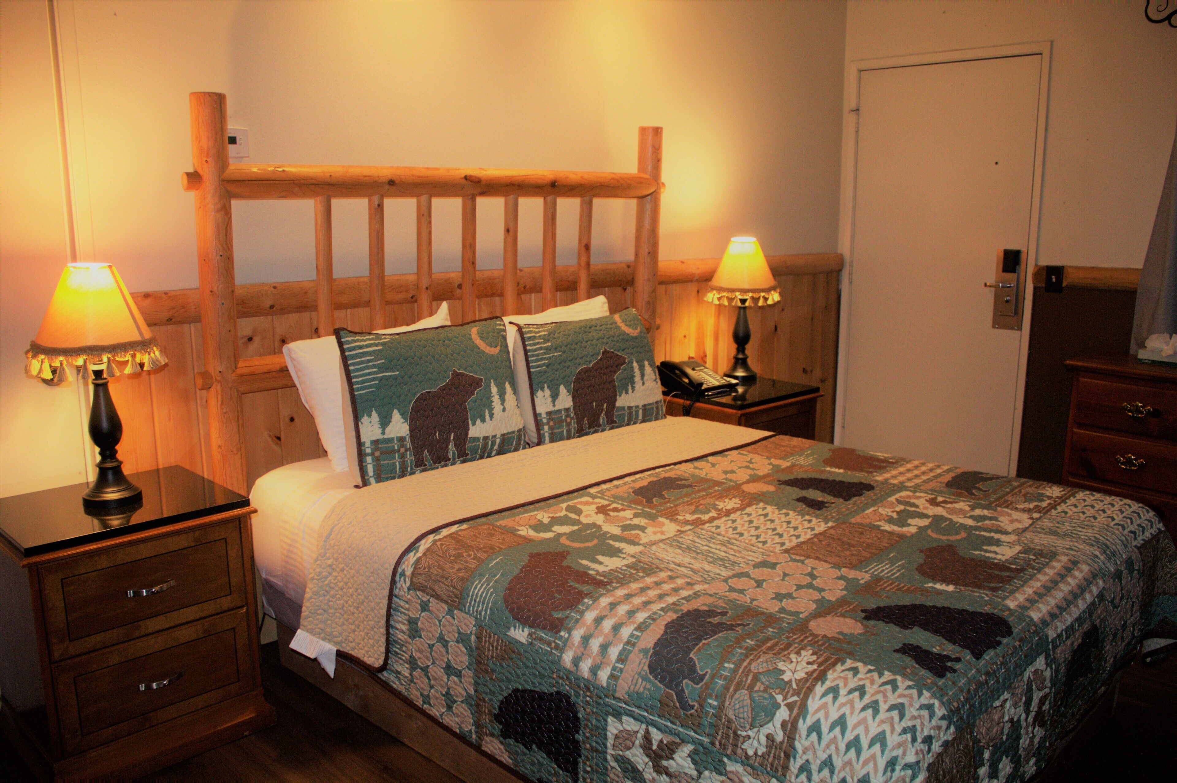 Quarto com cabeceira de madeira, colcha acolchoada, duas luminárias de cabeceira, mesa de cabeceira com gavetas e uma porta à direita.