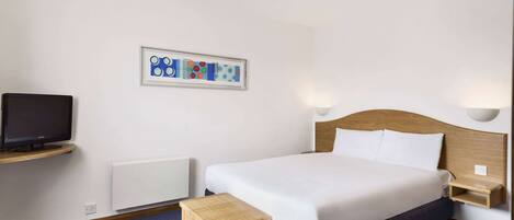 Quarto de hotel com cama, televisão sobre suporte, mesa de cabeceira, um pequeno banquinho de madeira e um quadro pendurado na parede.