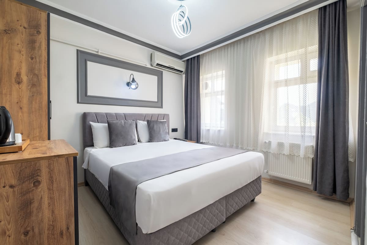 Quarto de hotel moderno com uma cama grande, uma mesa de cabeceira de madeira, um quadro emoldurado acima da cama e uma janela com cortinas.